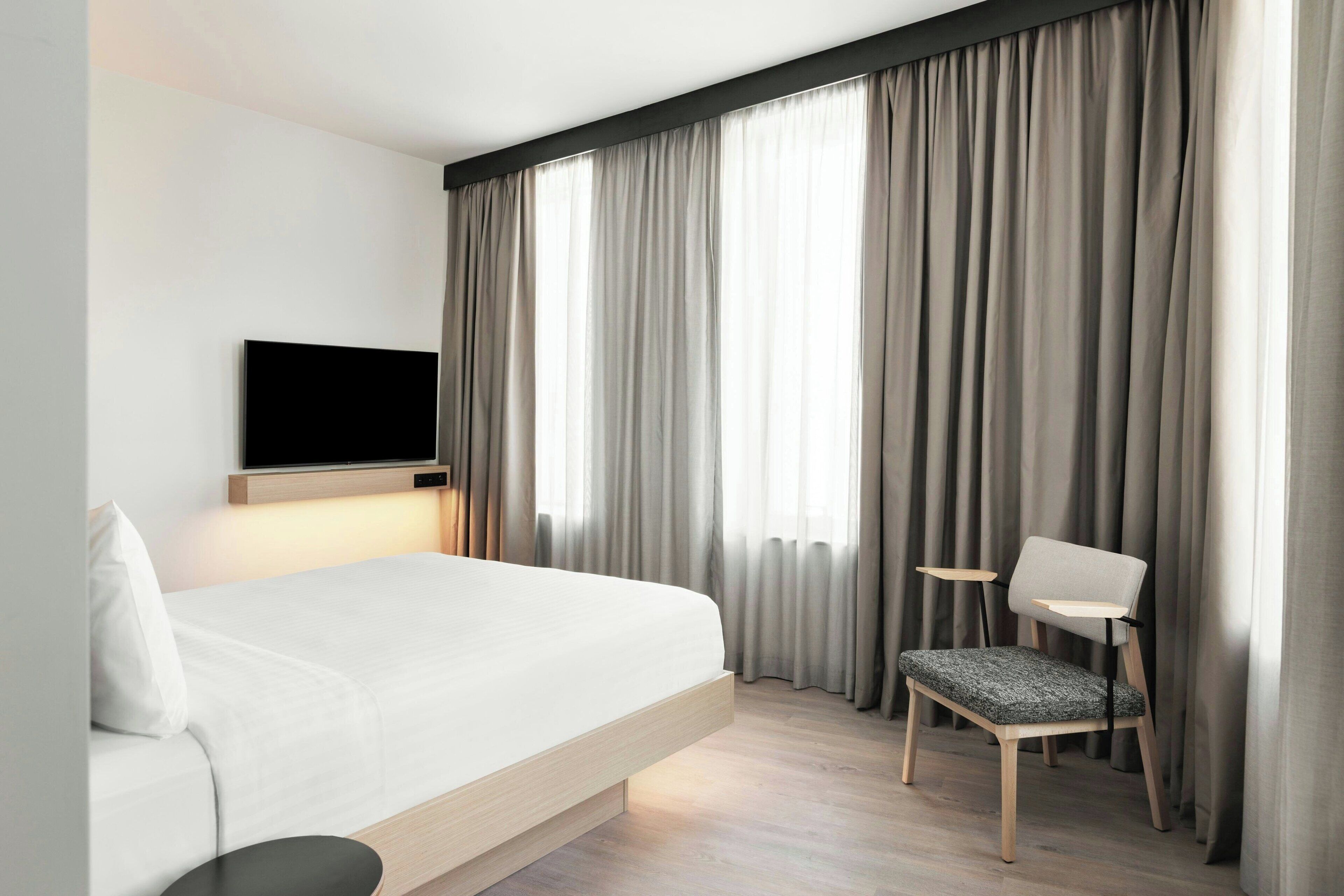 Quarto de hotel moderno com uma cama grande, televisão de tela plana na parede, uma mesinha e uma cadeira.