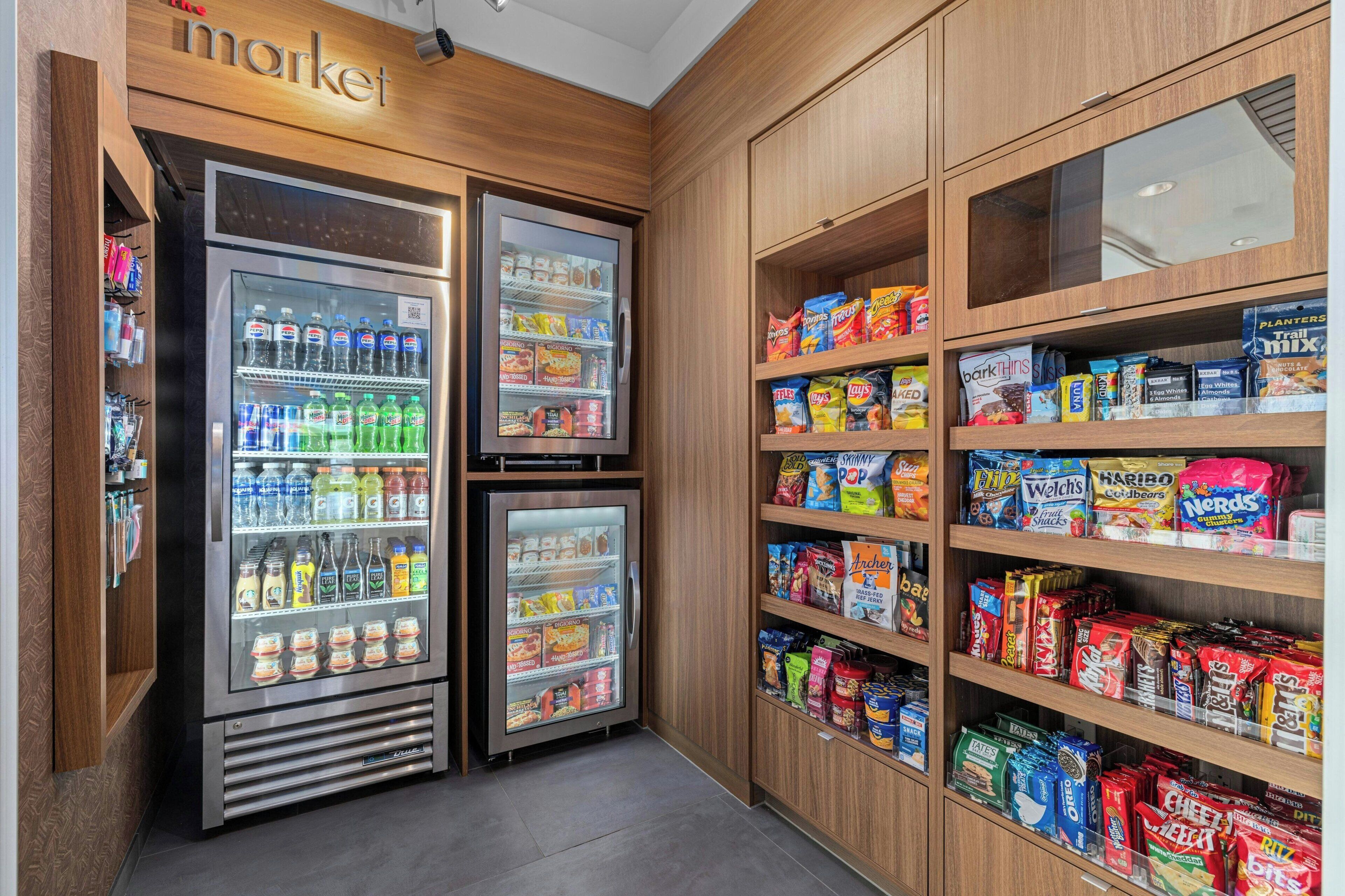 Uma área de conveniência com bebidas geladas em dispensadores e prateleiras repletas de lanches e cereais.