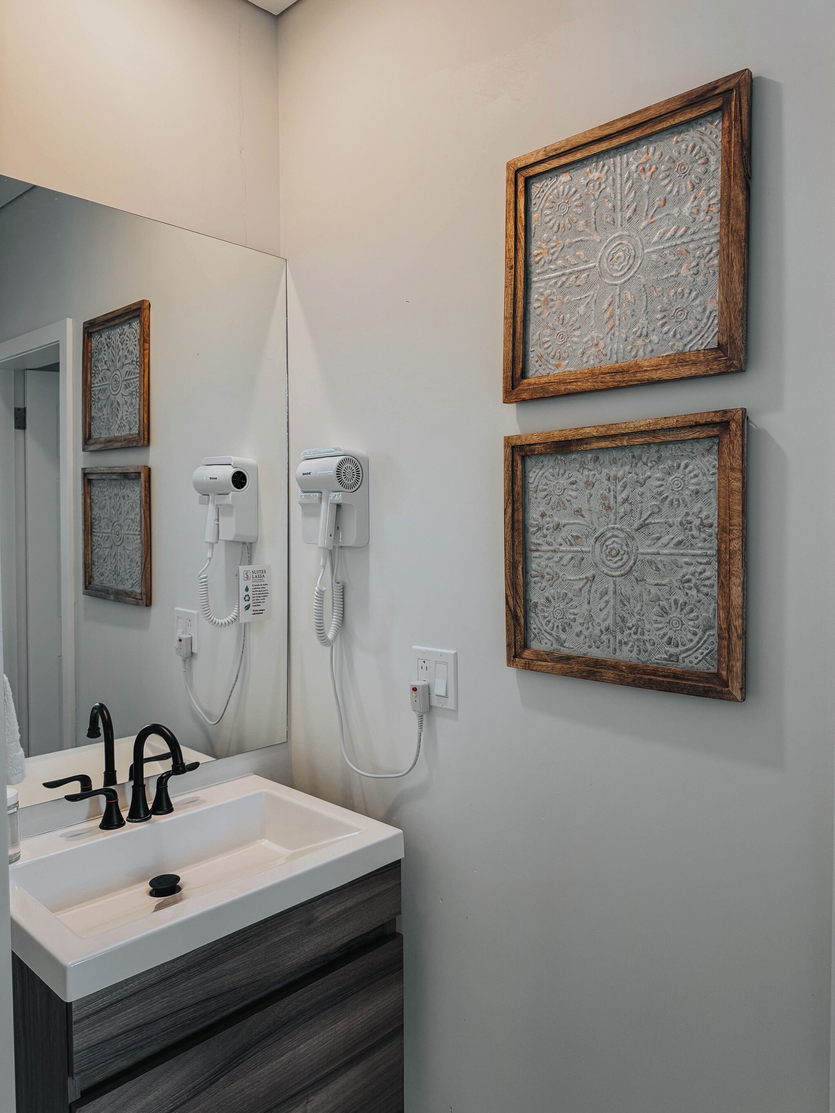 Casa de banho com lavatório branco, torneira preta e secador de cabelo fixado na parede. Existem duas decorações emolduradas sobre o lavatório.