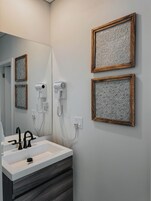 Casa de banho com lavatório branco, torneira preta e secador de cabelo fixado na parede. Existem duas decorações emolduradas sobre o lavatório.