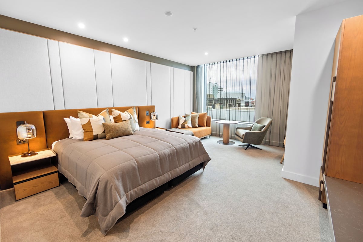 Quarto de hotel moderno com uma cama grande, um sofá, uma mesinha e vista da cidade através de janelas amplas.