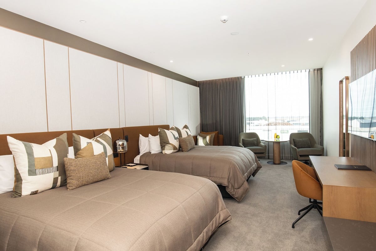 Quarto de hotel com duas camas, uma escrivaninha, uma cadeira, uma mesinha e uma janela grande.