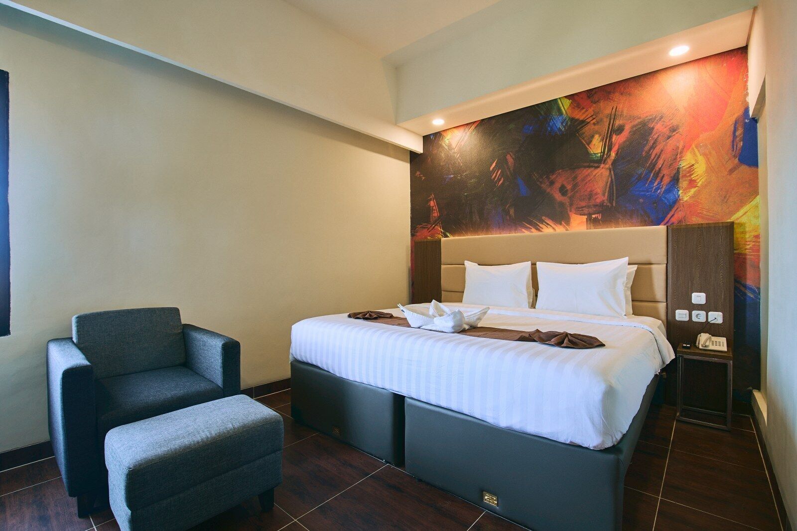Quarto de hotel com uma cama, uma cadeira, uma mesinha de cabeceira e uma pintura abstrata colorida na parede.