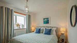 Quarto com cama, duas luminárias de cabeceira, janela com cortinas, um quadro na parede e um espelho.