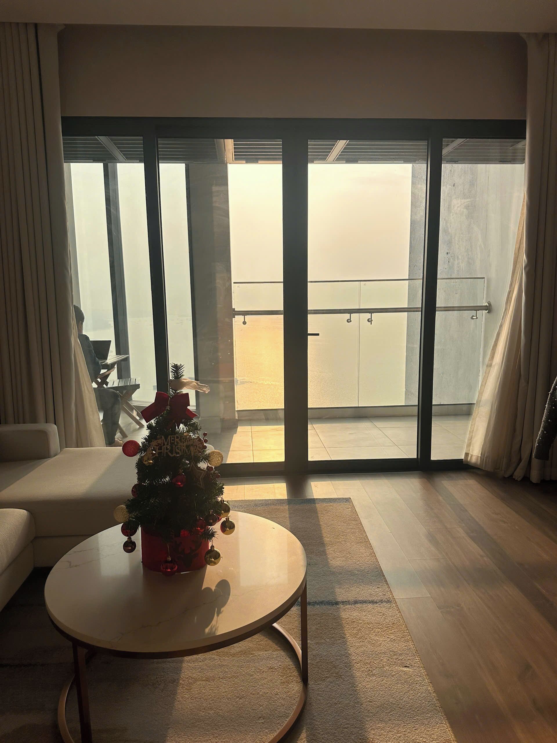 Sala moderna com árvore de Natal sobre uma mesa redonda, grandes portas de vidro deslizantes para uma varanda e um sofá branco.