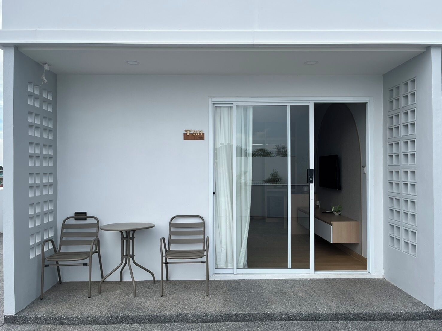 Uma pequena área externa com dois cadeiras e uma mesa, uma porta de vidro deslizante que dá para uma varanda, e uma pequena placa acima da porta.