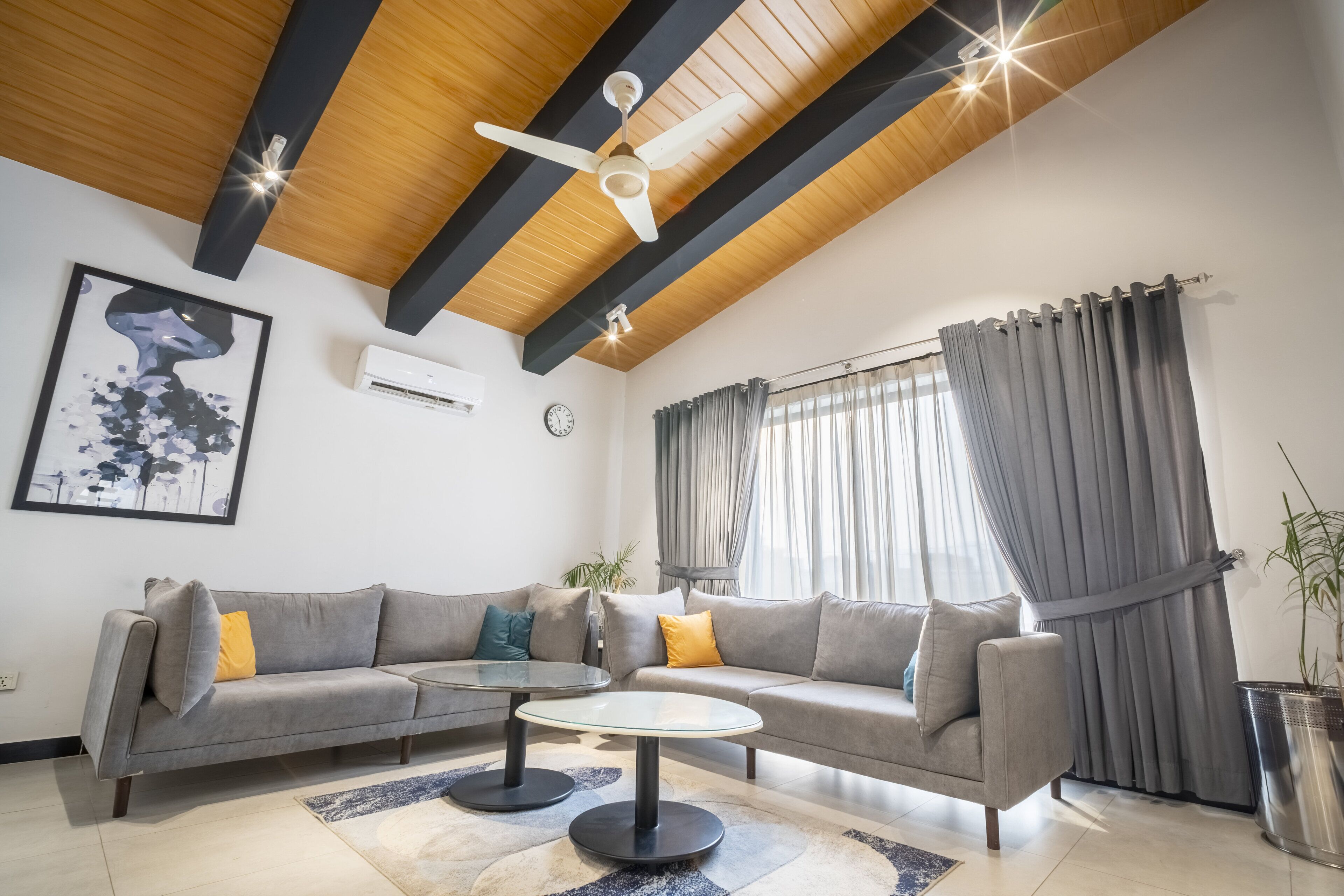 Uma sala de estar moderna com um sofá cinzento, uma mesa de centro de vidro, um ventilador de teto e uma janela grande com cortinas cinzas.