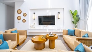 Uma sala de estar moderna com uma TV de tela plana na parede, duas mesas de centro de madeira, sofás bege com almofadas azuis e uma grande planta em vaso.