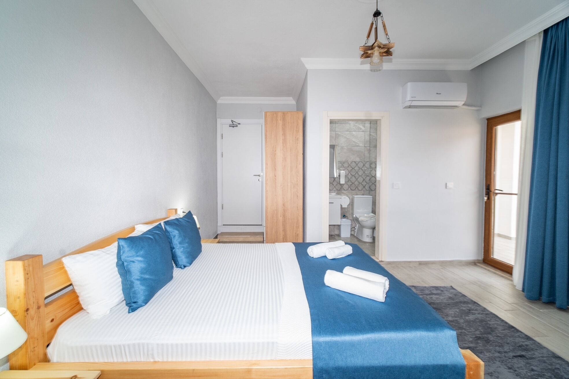 Quarto com cama, almofadas, mesa de cabeceira, luminária pendente, banheiro com chuveiro e ar condicionado.