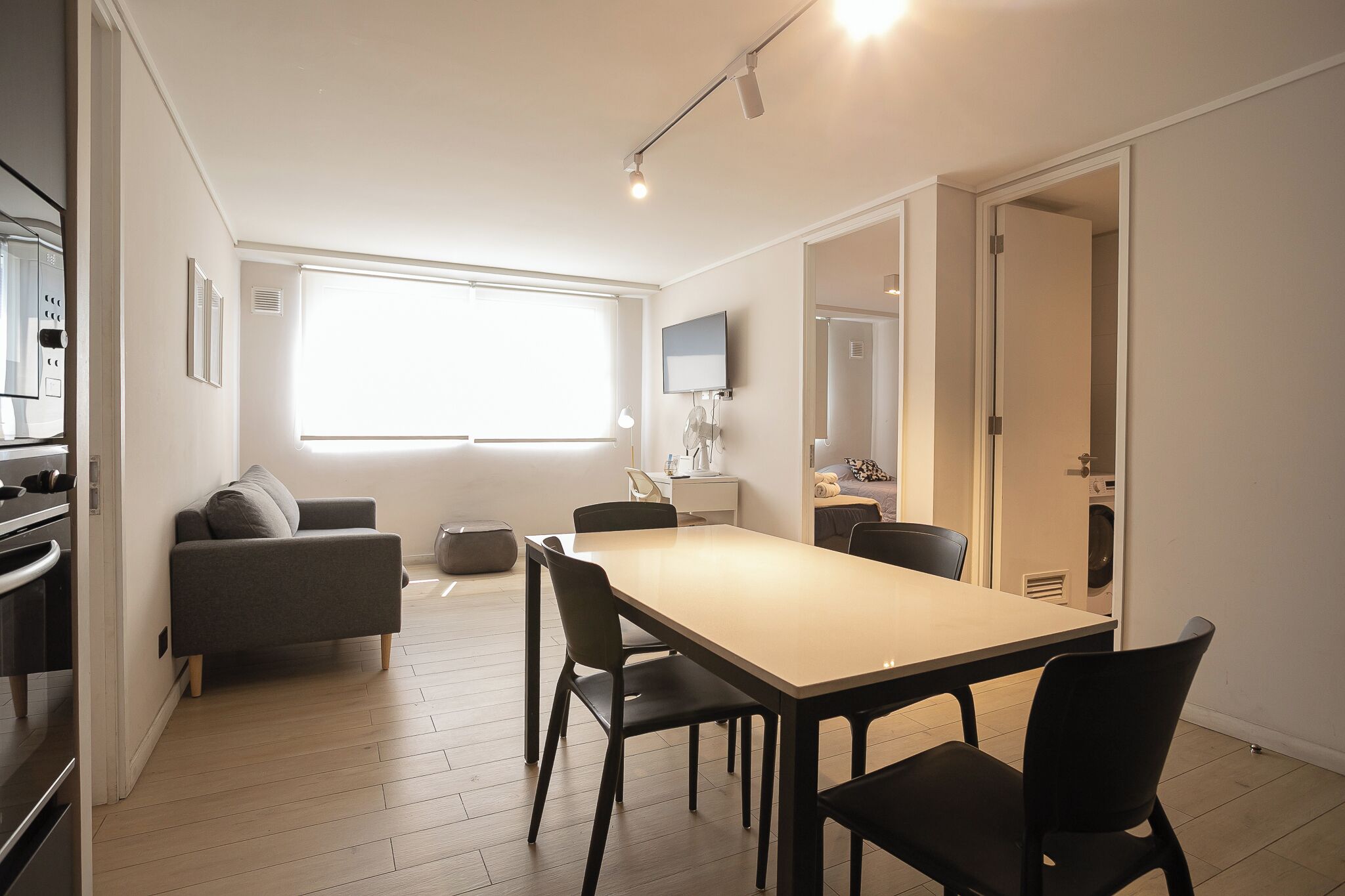 Um quarto moderno e bem iluminado com uma mesa de jantar, cadeiras, um sofá e uma televisão fixada na parede.