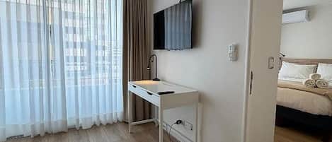 Quarto de hotel com piso de madeira, uma escrivaninha com cadeira, uma cama e uma televisão fixada na parede.