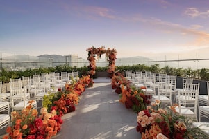 Um espaço para eventos ao ar livre com um arco floral, cadeiras brancas e vista desobstruída da paisagem urbana ao fundo.
