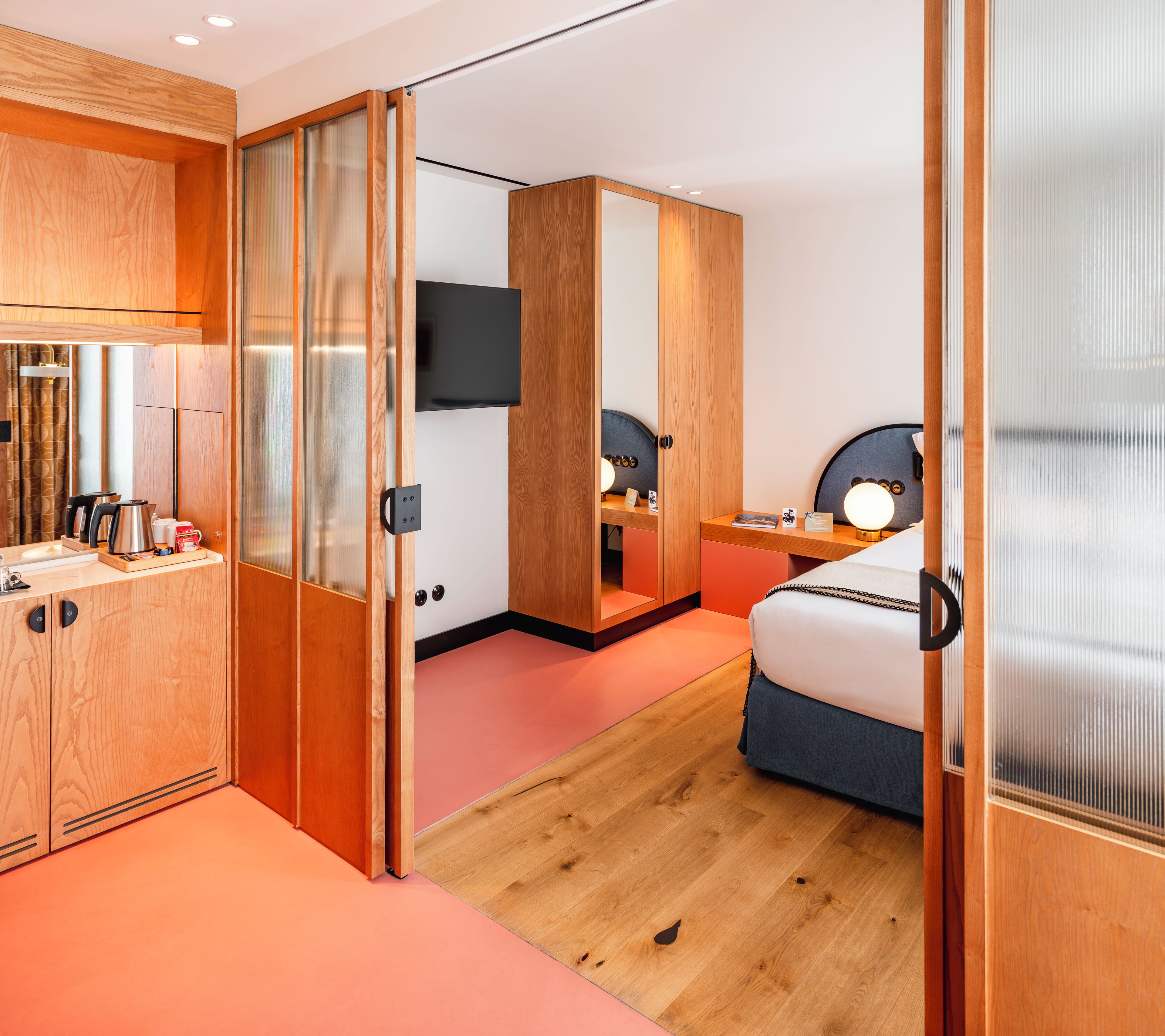 Quarto de hotel moderno com armário de madeira, cama com cabeceira, mesa de cabeceira com luminária e televisão de tela plana fixada na parede.
