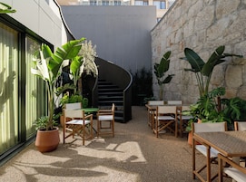 Área externa com mesas e cadeiras, rodeada por plantas em vasos e uma parede de pedra.
