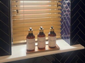 Três frascos de gel de banho e condicionador numa prateleira em uma casa de banho com persianas de madeira e parede com padrão azul.
