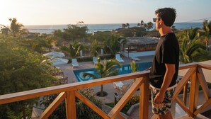 Um homem está em um deck de madeira com vista para uma área de piscina com espreguiçadeiras e palmeiras.