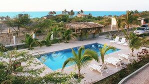 Área da piscina com espreguiçadeiras, estrutura com telhado de palha e vista para o oceano.