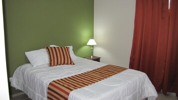 Cama bem arrumada com lençóis brancos e colcha listrada, abajur e cortina vermelha.
