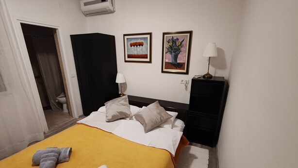 Quarto de hotel com uma cama, duas almofadas, um abajur, cômoda e dois quadros na parede.