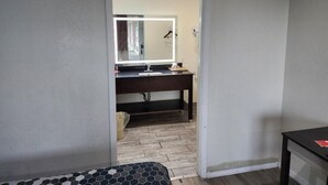Quarto com piso de madeira, escrivaninha escura, espelho e porta branca que dá para outro cômodo.