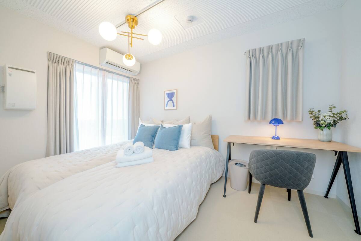 Quarto de hotel com cama, escrivaninha, cadeira, luminária e janela com cortinas.