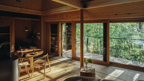 Interior moderno de cabana com piso de madeira, área de refeições com cadeiras, cozinha com fogão e grandes janelas com vista para a floresta.