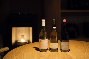 Três garrafas de vinho sobre uma mesa redonda de madeira, com uma lareira acesa ao fundo.