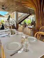 Um restaurante com mesas brancas, cadeiras de vime e uma escadaria que dá para uma área externa.