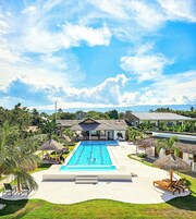 Piscina rodeada por construções com telhados de palha, uma área gramada e vista para montanhas ao longe.