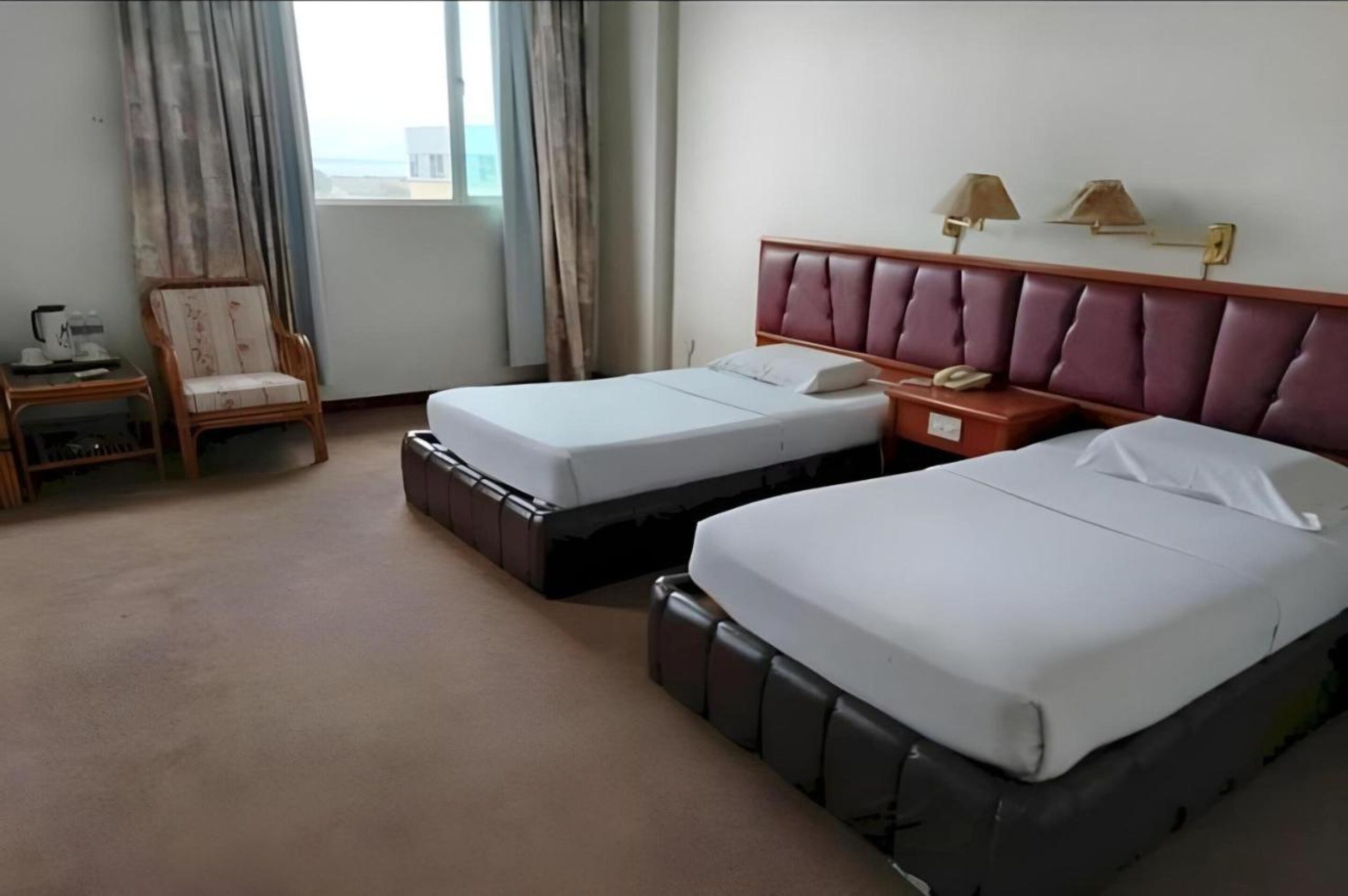 Quarto de hotel com duas camas, uma cadeira, uma mesa de cabeceira, um abajur e uma janela com cortinas.