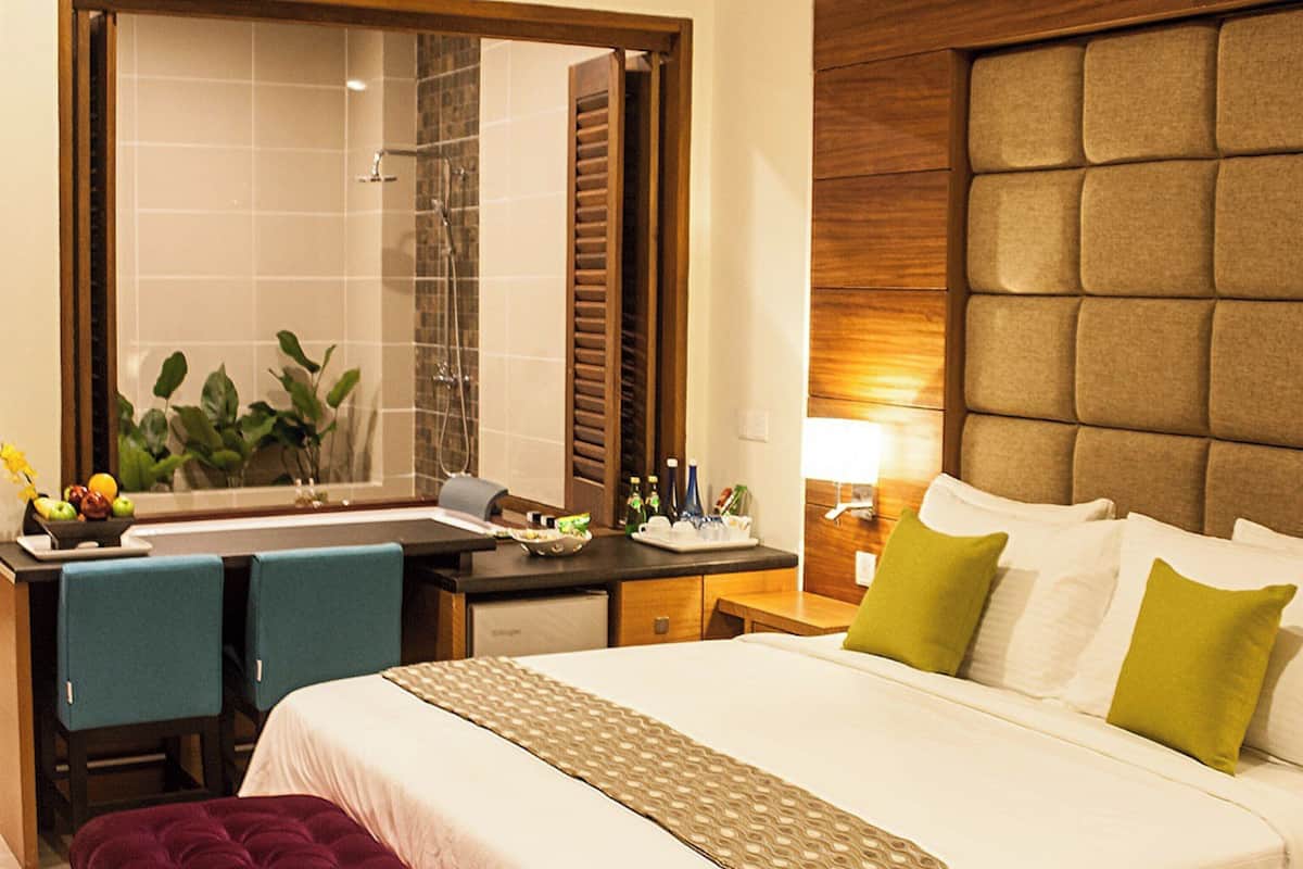 Quarto de hotel com uma cama grande, uma mesa com duas cadeiras, um espelho e uma cabeceira de madeira.