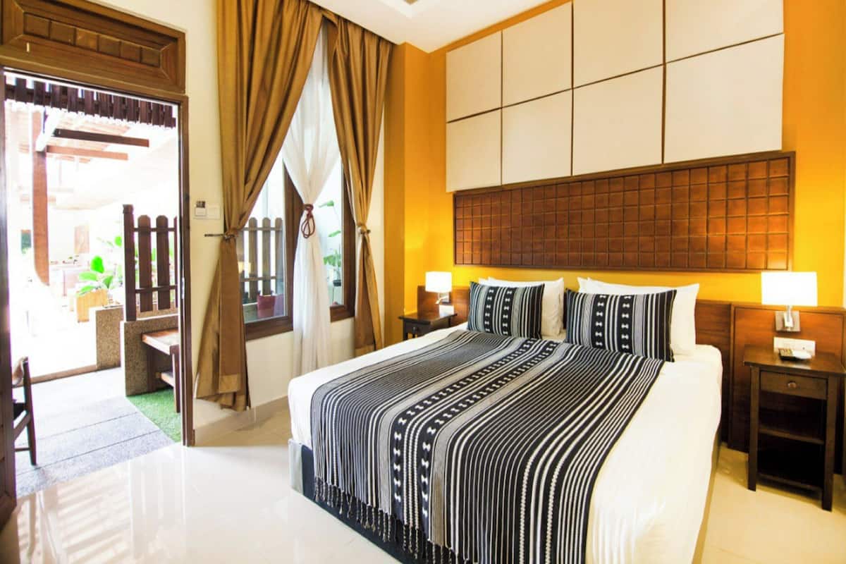 Quarto de hotel com uma cama grande, duas mesas de cabeceira com luminárias, uma cabeceira de madeira e vista para o exterior através de uma janela com cortinas.