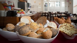 Diversos pães e doces sobre uma mesa em uma sala de jantar, com cozinha ao fundo.