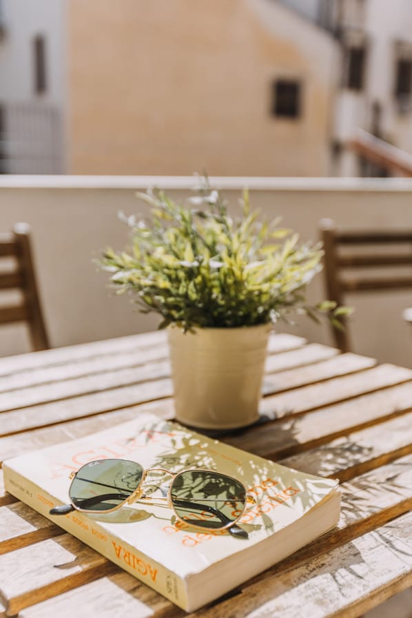 Um livro intitulado "Acqua Alta" com óculos de sol em cima, colocado sobre uma mesa de madeira com uma planta em vaso ao lado.