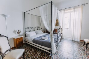 Uma cama com dossel e cortinas, mesa de cabeceira com abajur, uma cadeira e piso estampado.
