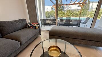 Sala de estar moderna com sofá cinzento, mesa de centro de vidro e vista para uma piscina e edifícios.
