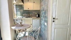 Uma cozinha compacta com pia, fogão e um pequeno frigorífico. Há uma mesa redonda com um objeto decorativo e uma porta à direita.