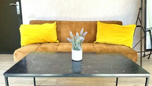 Quarto com sofá marrom, duas almofadas amarelas, mesa de centro de mármore preto e um vaso branco com planta.