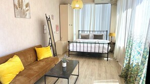 Quarto com cama, mesa de cabeceira, abajur, sofá com almofadas, pequena mesa com um vaso e janela com cortinas.
