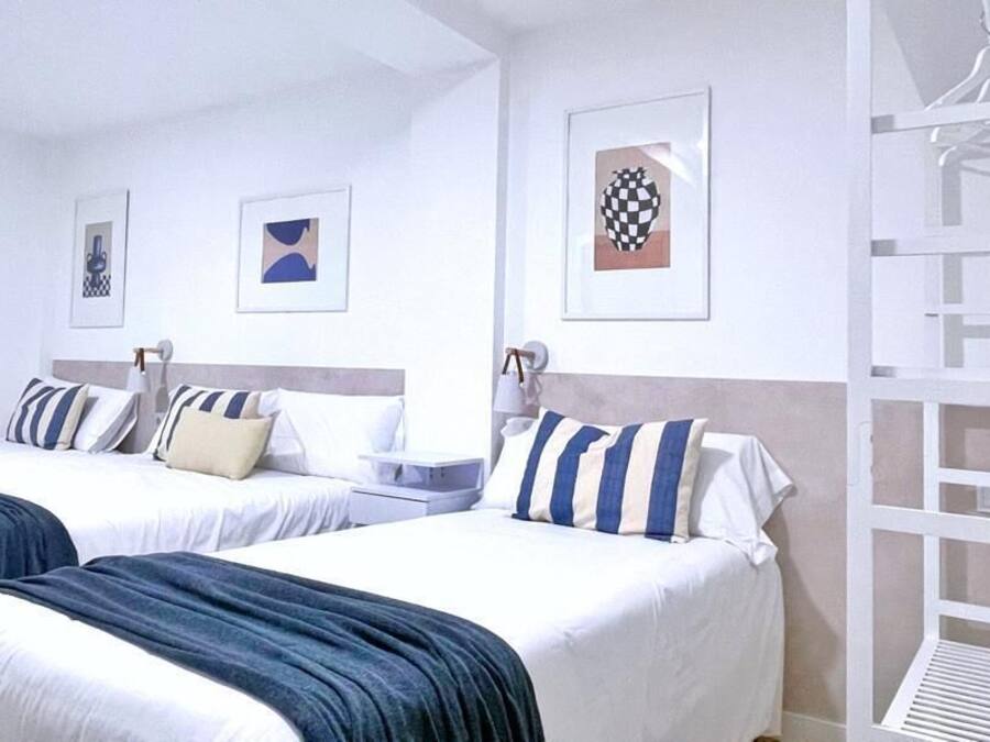 Quarto de hotel com duas camas, cada uma com almofada listrada e manta azul. Há quadros nas paredes e uma estante branca.