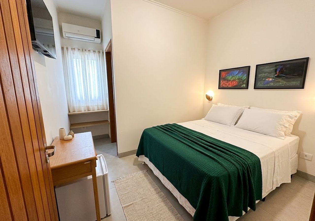 Quarto pequeno e bem iluminado, com uma cama, uma escrivaninha e uma janela com cortinas.
