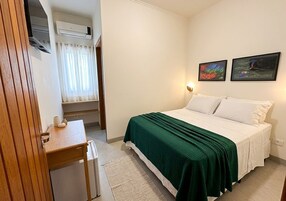 Quarto pequeno e bem iluminado, com uma cama, uma escrivaninha e uma janela com cortinas.