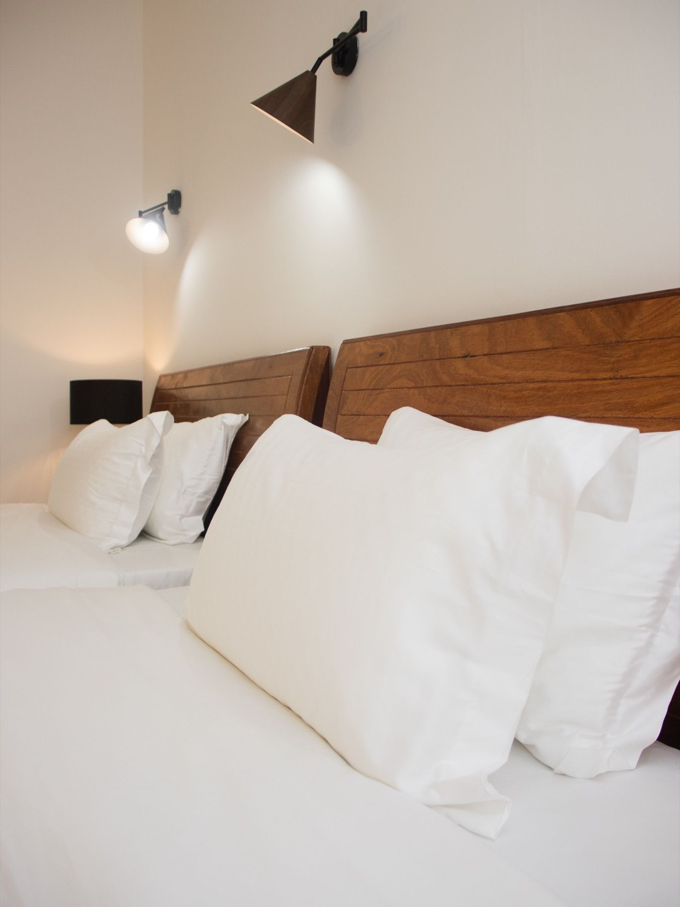 Quarto de hotel com cabeceira de madeira, roupa de cama branca e luminária fixada na parede.