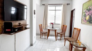 Uma sala de estar moderna com uma televisão de tela plana presa a uma prateleira de madeira, uma mesa de jantar redonda com quatro cadeiras, uma mesinha lateral com um copo e um quadro na parede.