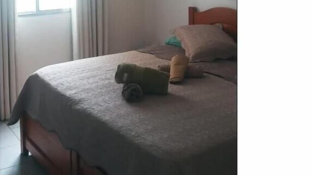 Quarto com cama, ventoinha no teto, janela com cortinas e mesa de cabeceira.