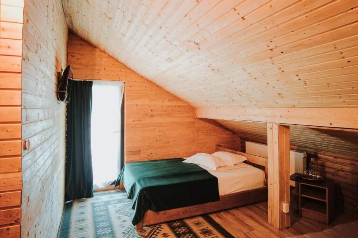 Um quarto pequeno e aconchegante com paredes de madeira e teto inclinado. Possui uma cama de solteiro com cobertor verde e almofadas brancas, uma mesa pequena com abajur e uma janela com cortina.