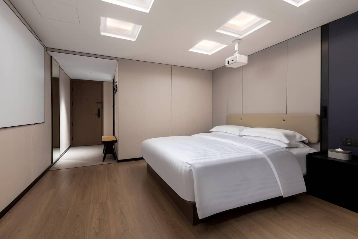 Quarto de hotel moderno com uma cama grande, uma mesa de cabeceira, um projetor no teto e piso de madeira.
