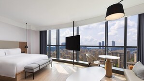 Quarto de hotel moderno com uma cama grande, uma escrivaninha, uma cadeira, uma mesa redonda e uma televisão. O quarto possui amplas janelas com vista para a cidade.