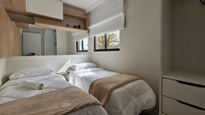 Quarto moderno com duas camas, um guarda-roupa embutido e uma janela com vista para árvores.