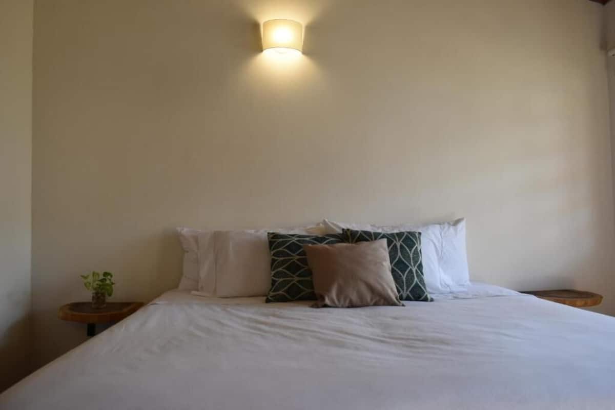 Cama bem arrumada com roupa de cama branca e várias almofadas, incluindo uma com padrão verde. Há uma pequena planta sobre uma mesinha de cabeceira de madeira à esquerda.